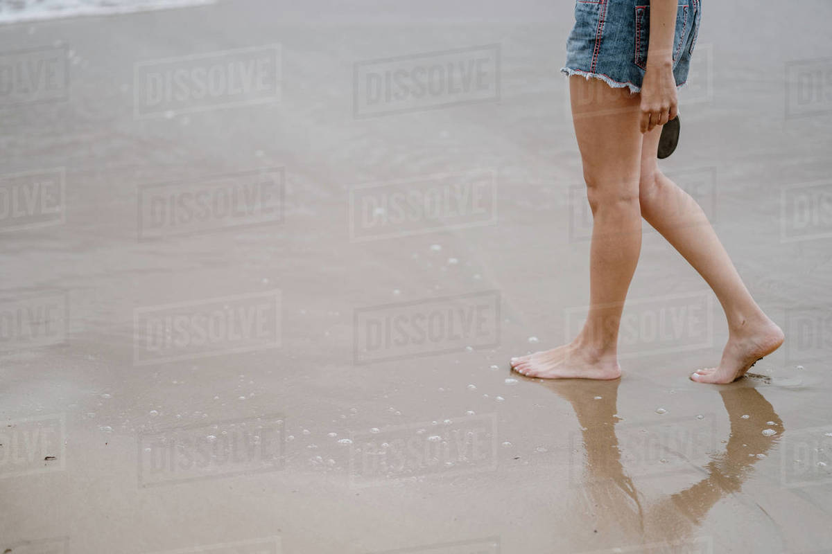 Side view of crop woman in jean shorts walking with shoes in hand on ...