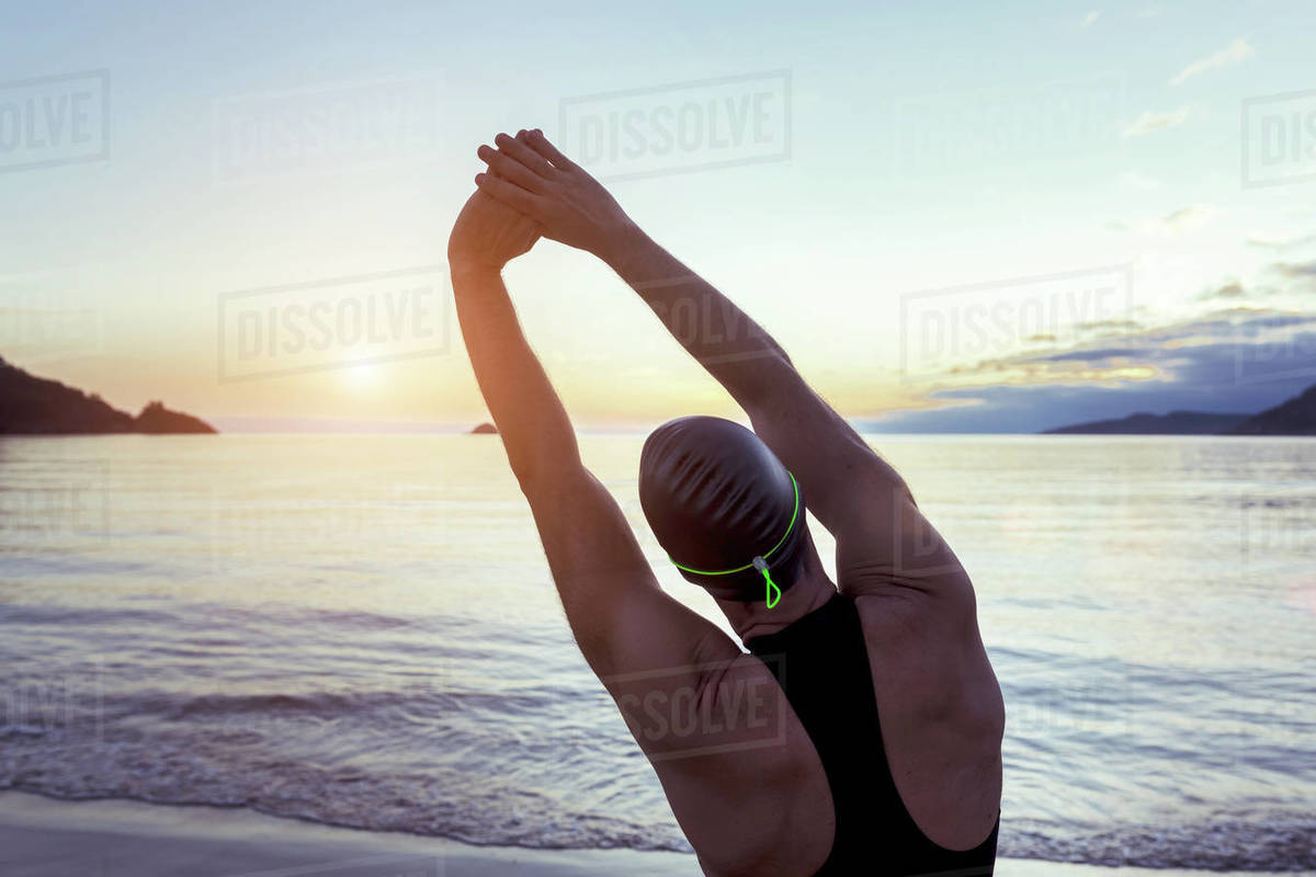 Back view of fit male swimmer in swimsuit and cap standing on seashore ...