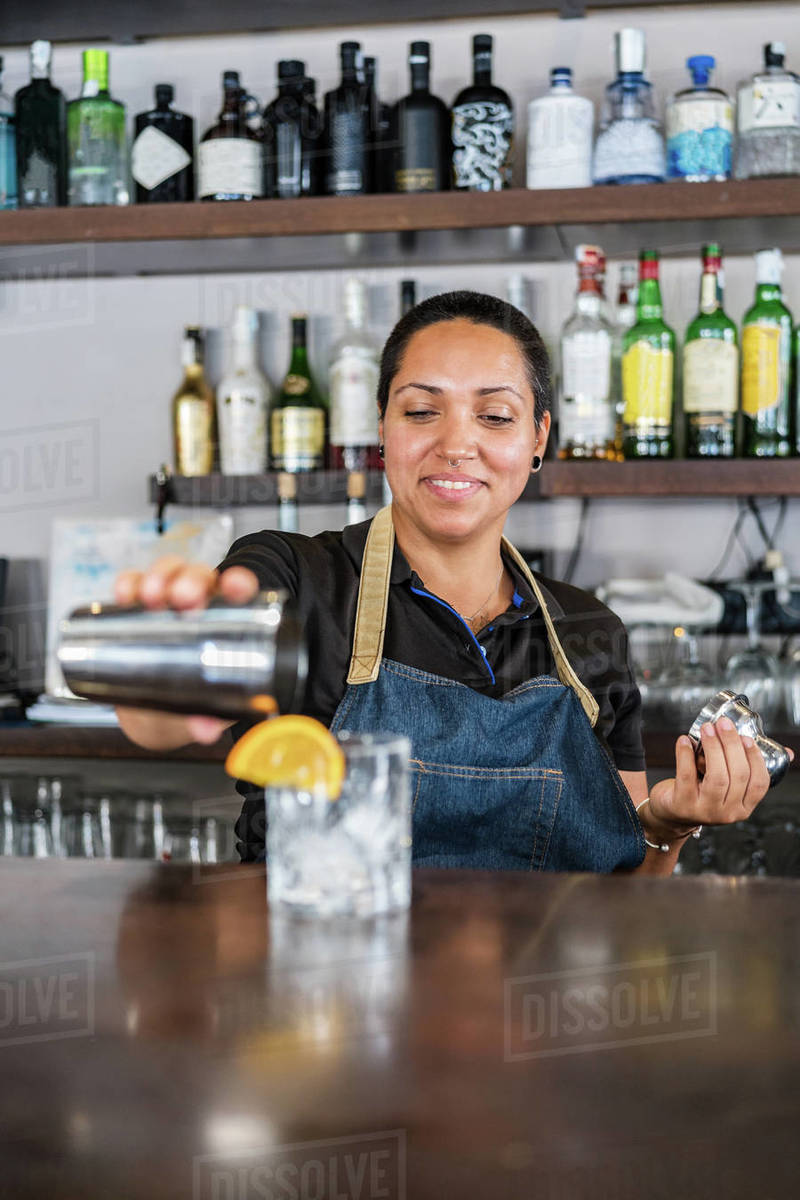 Positive focused female barkeeper in apron standing at counter making ...