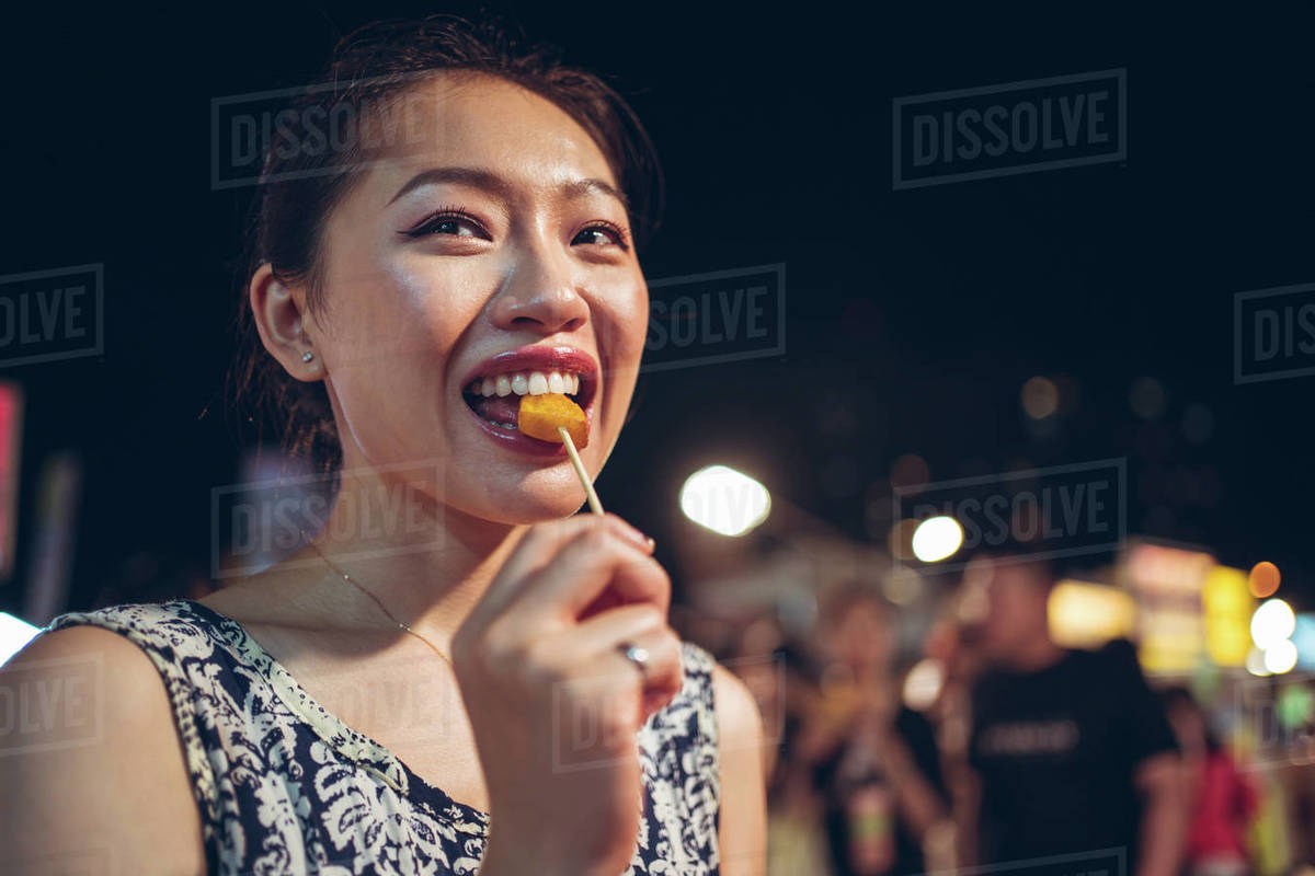 Delighted Asian female eating delicious sugar coated potato on stick ...