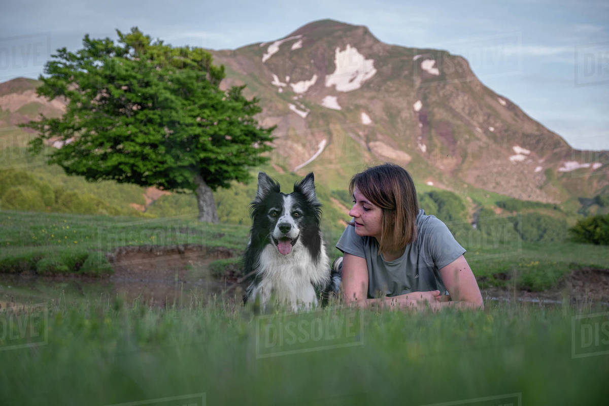 A girl and her border collie dog lying in the grass with a tree and ...