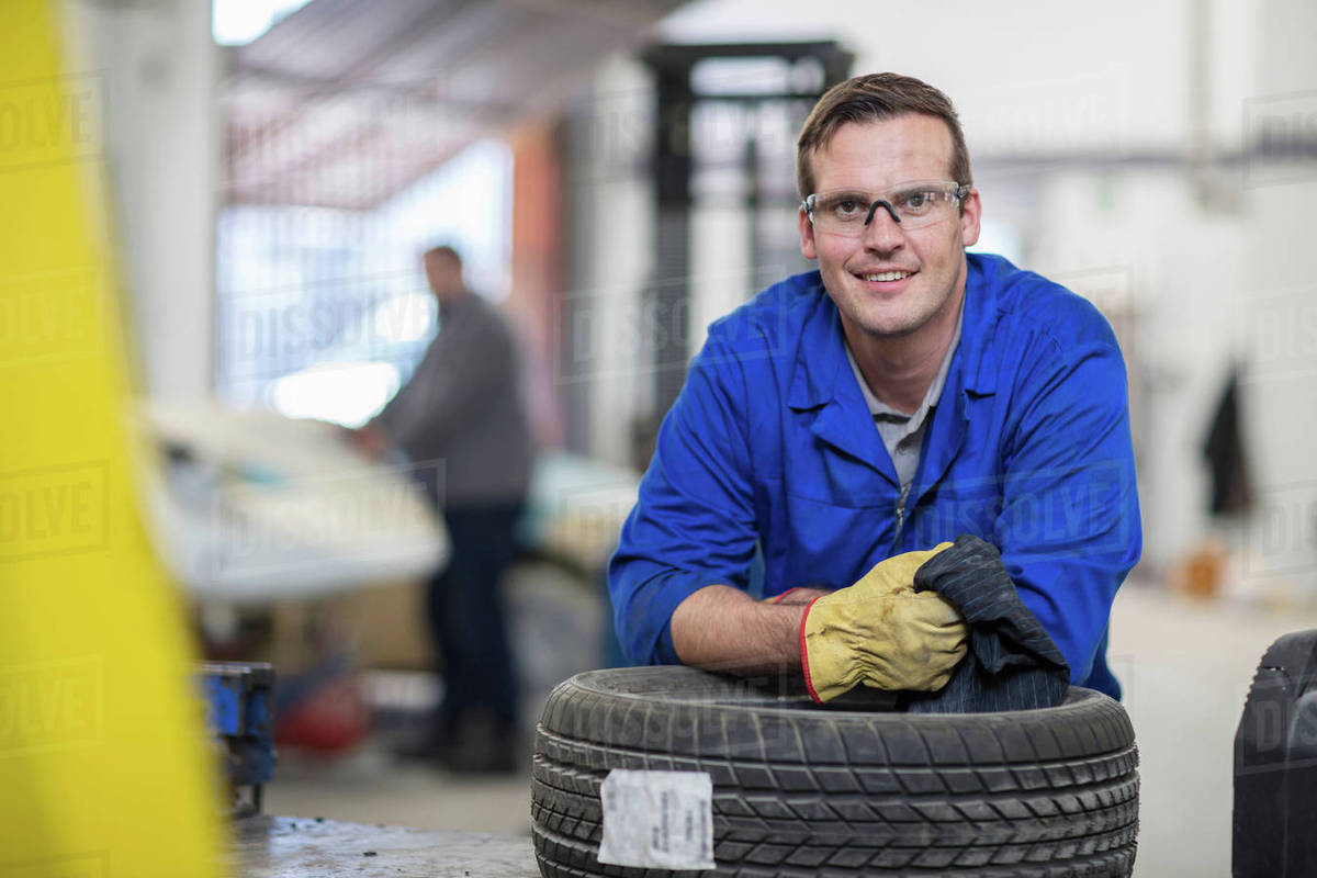 Portrait of male car mechanic leaning against tyres in repair garage ...