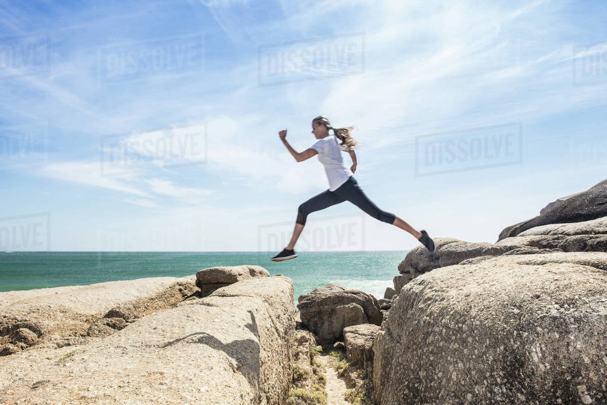 Young female runner leaping over beach rocks, full length, Cape Town ...