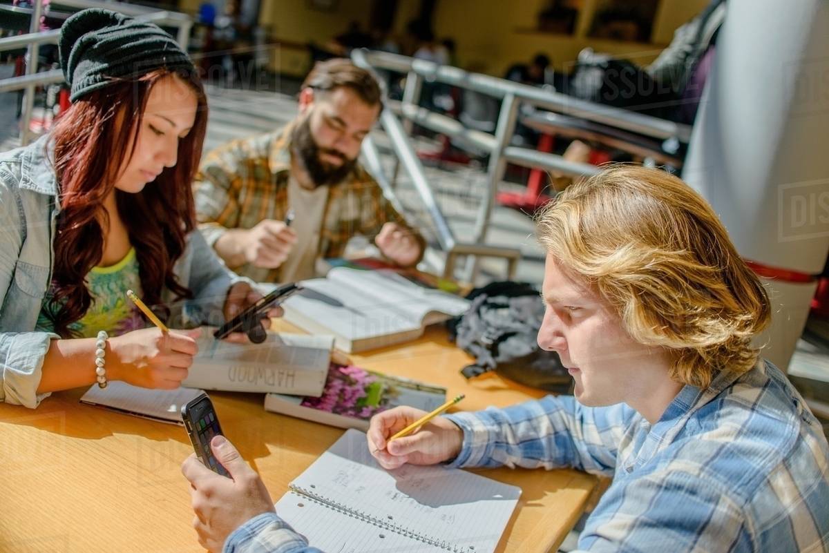 Three adult college students making notes and using smartphones at ...