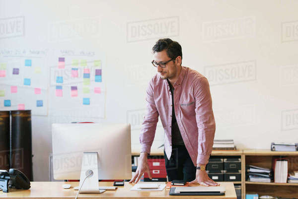 Man in office leaning against desk looking at computer monitor - Stock ...
