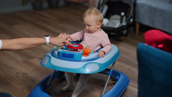 Concentrated adorable little girl sitting in baby walker as female hand ...