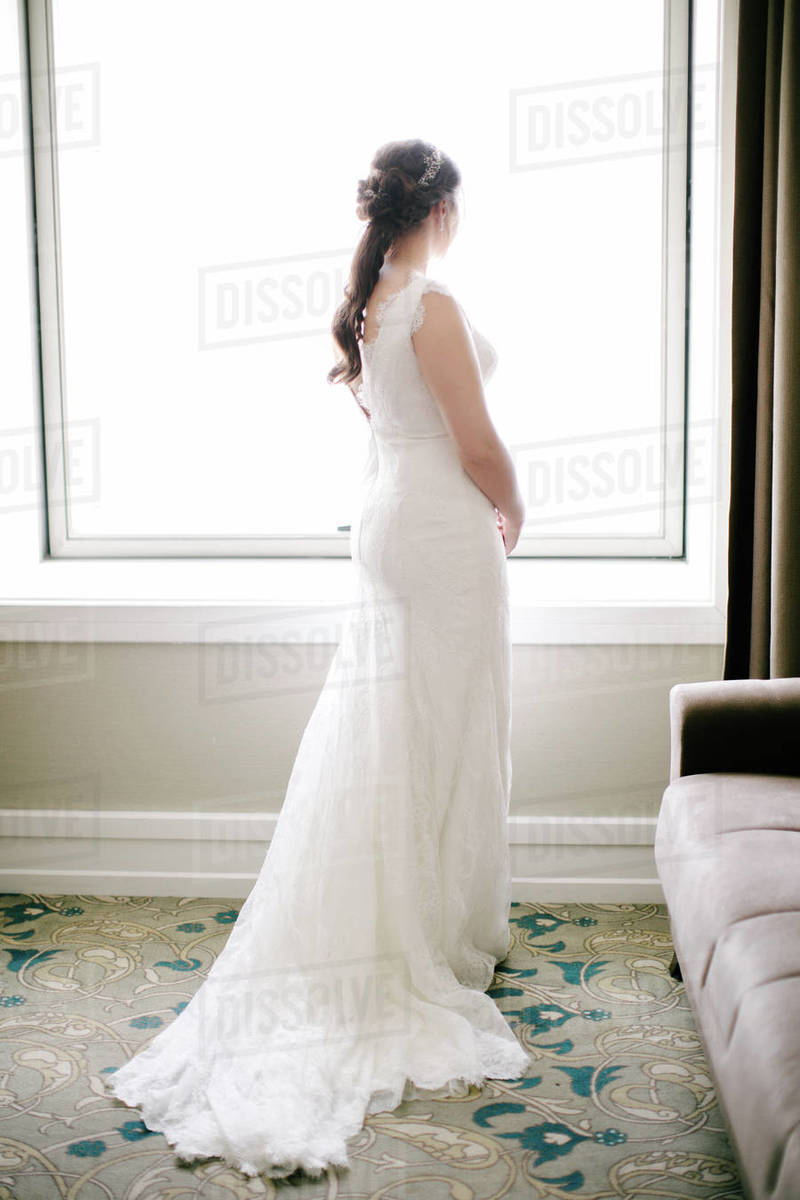 The bride poses facing back. She looks out the window. Indoor ...