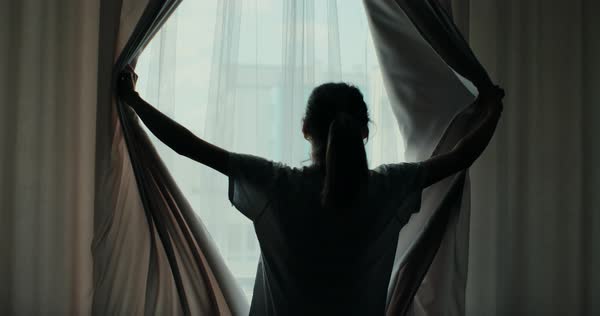 Young attractive woman approaches a window, and opens curtains. Warm ...