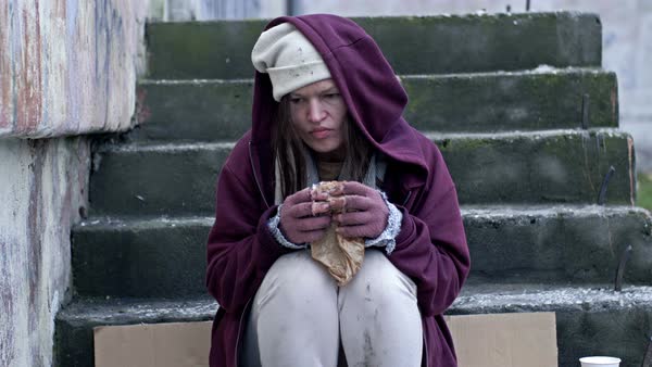 Homeless young woman, shivering with cold, greedily eats something ...