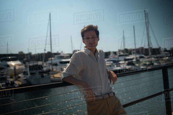 Portrait of a young man leaning on railings with boats moored at harbor ...