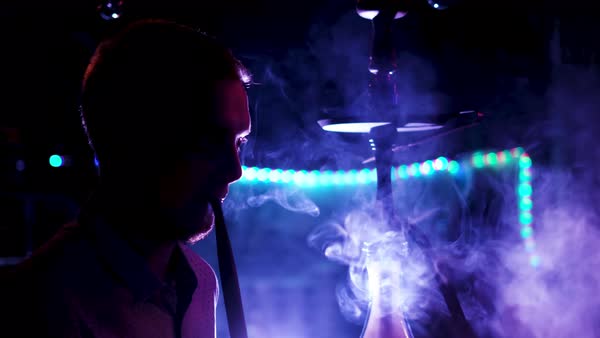 Side view of male silhouette in neon light smoking hookah, relaxing at ...