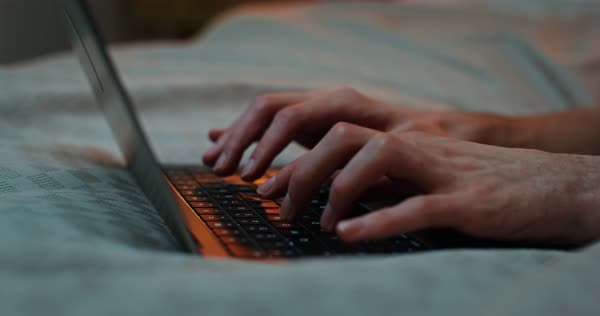 A man is typing on a laptop keyboard while lying on a bed in the ...