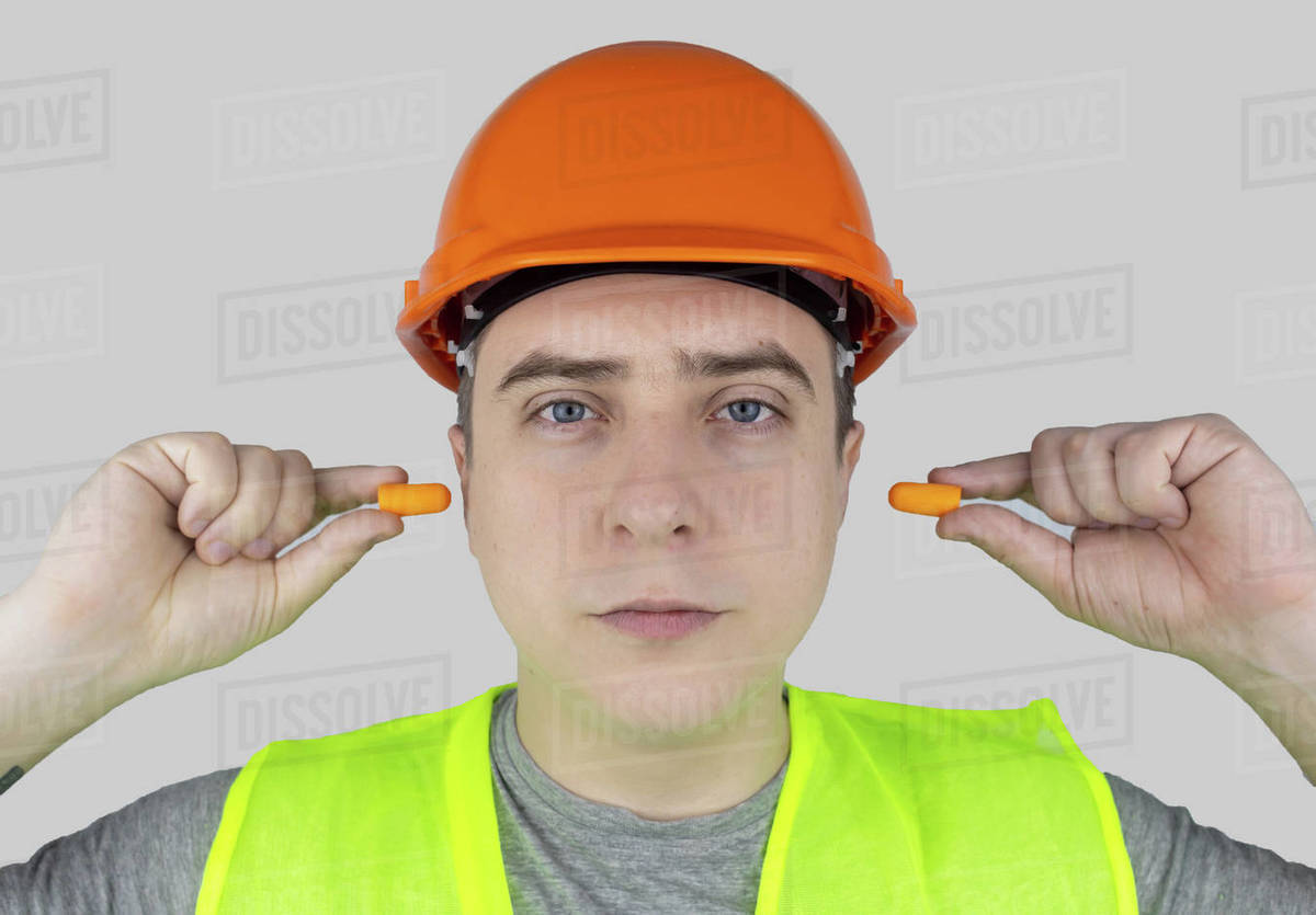A worker in a hard hat and green vest puts on ear plugs. Industrial ...