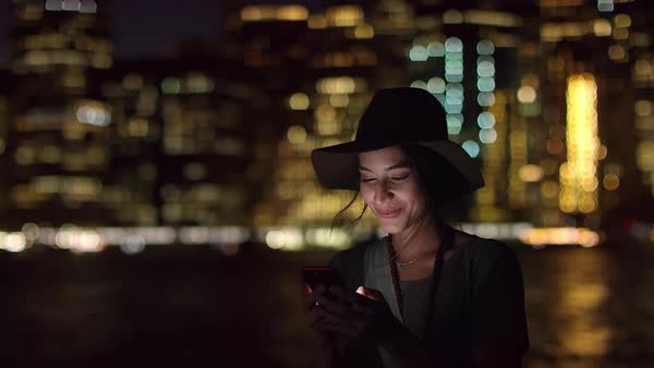 Woman uses mobile phone at night with city skyline in background ...