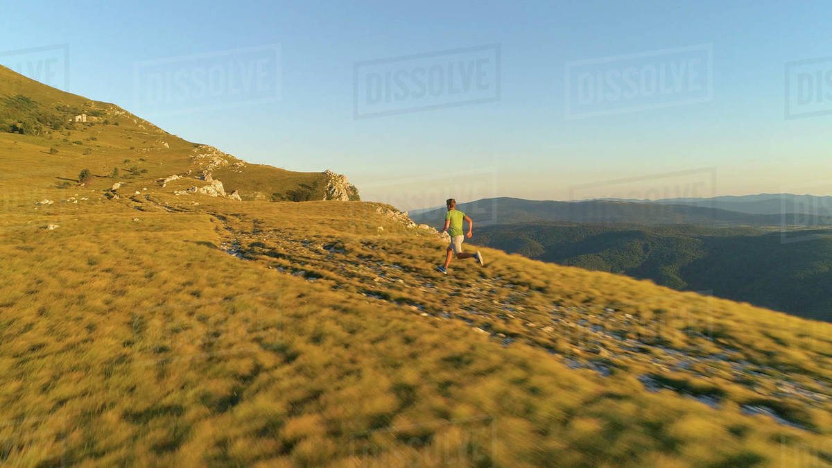 AERIAL: Flying behind an energetic man running up a grassy hill on a ...