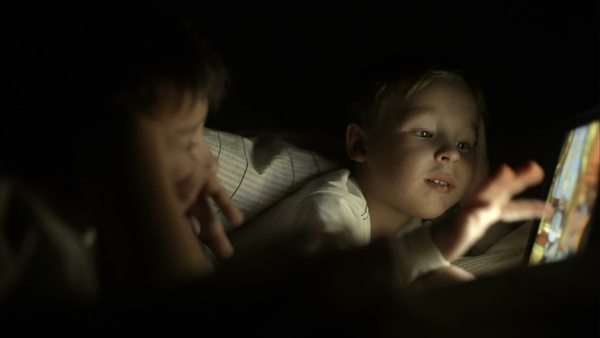 Two boys using tablet lying in bed at night. Bright screen illuminating ...