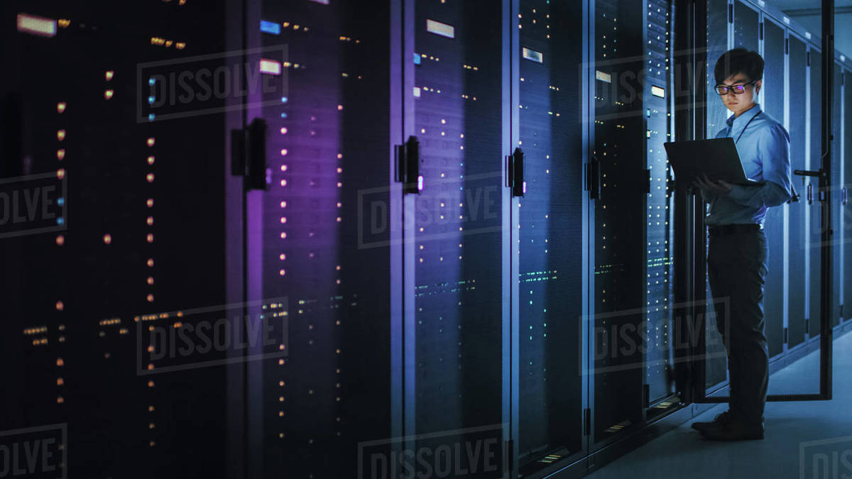 In Dark Data Center: Male IT Specialist Stands Beside the Row of Operational Server Racks, Uses ...