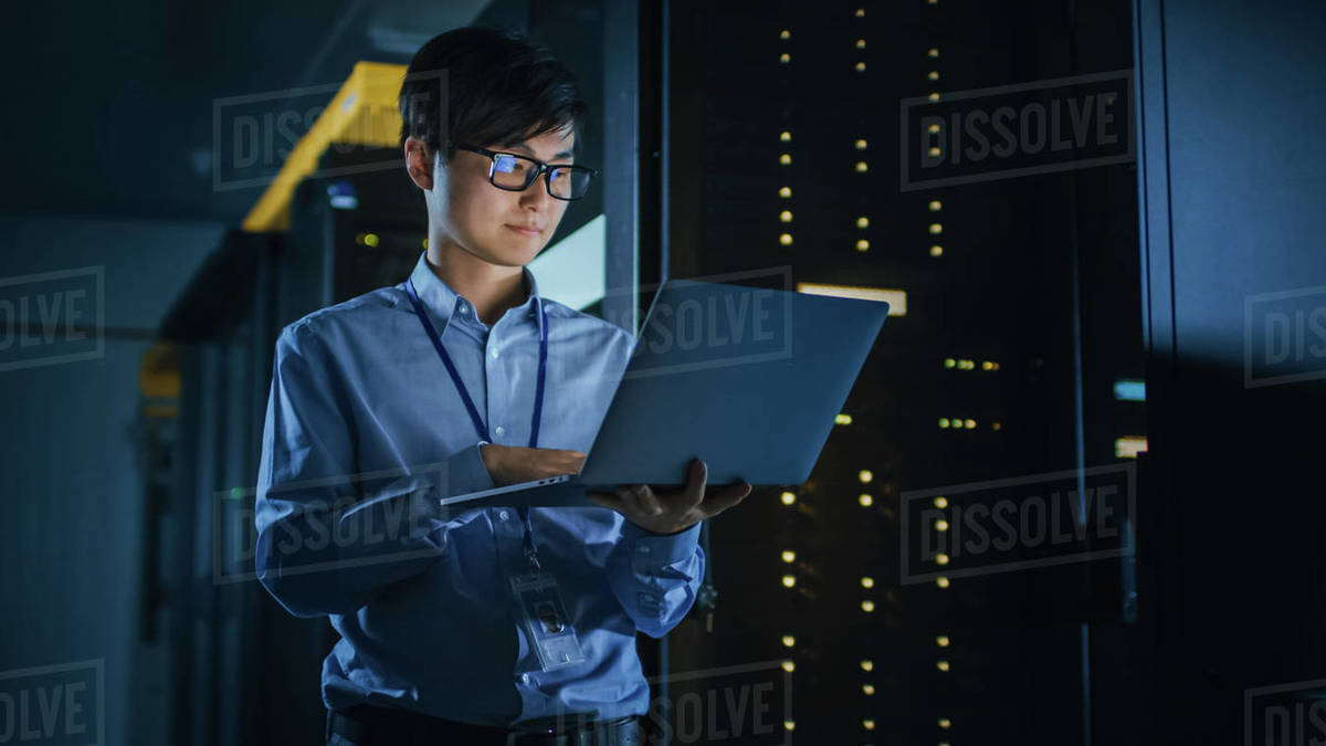 In Dark Data Center: Male IT Specialist Stands Beside the Row of Operational Server Racks, Uses ...