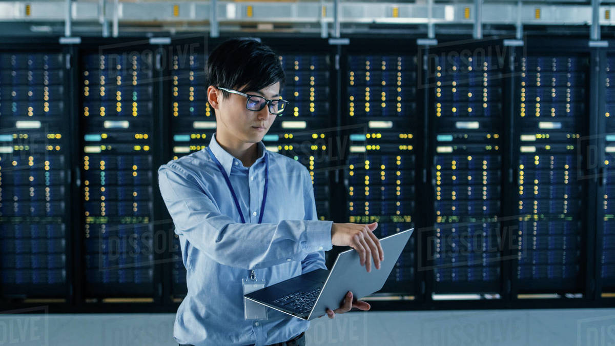 In Dark Data Center: Male IT Specialist Stands Beside Row of Operational Server Racks, Uses ...