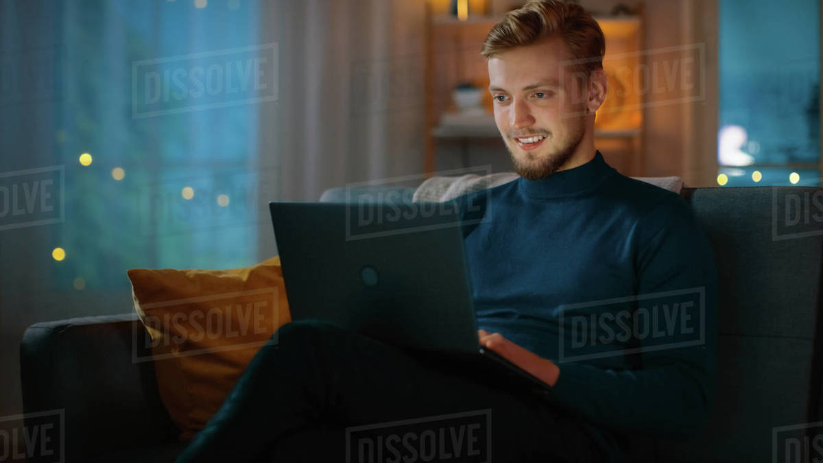 In the Evening Handsome Young Man at Home Sitting on a Couch Works on a ...