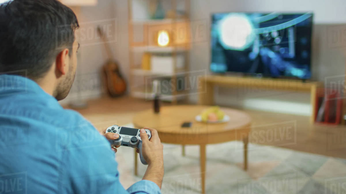 In the Living Room Man Sitting on a Couch Holds Controller Playing in a ...