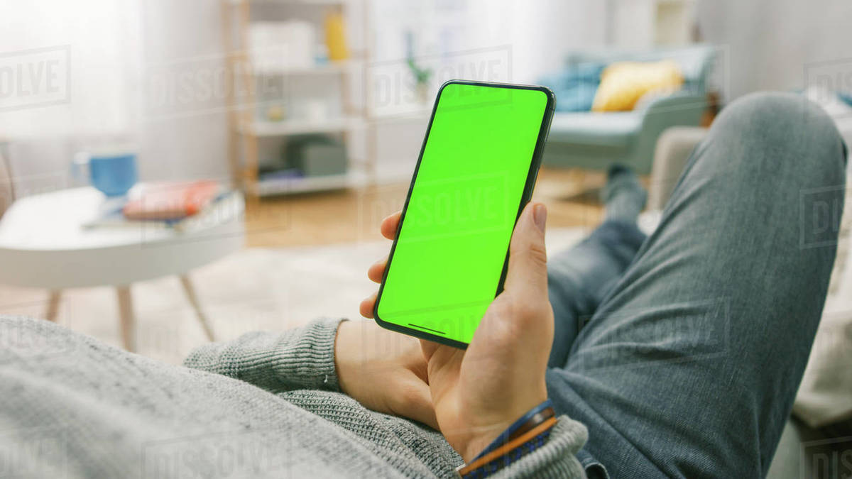 Man at Home Lying on a Couch using Smartphone with Green Mock-up Screen. Over the Shoulder ...