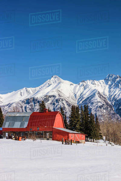A red roofed barn in a snow-covered field with the Chugach Mountains in ...