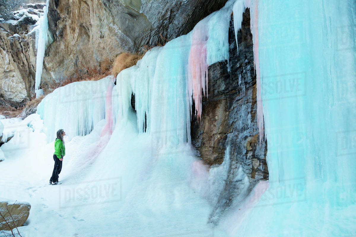 A woman hiker looks up at a frozen ice fall of pink and blue ice in ...