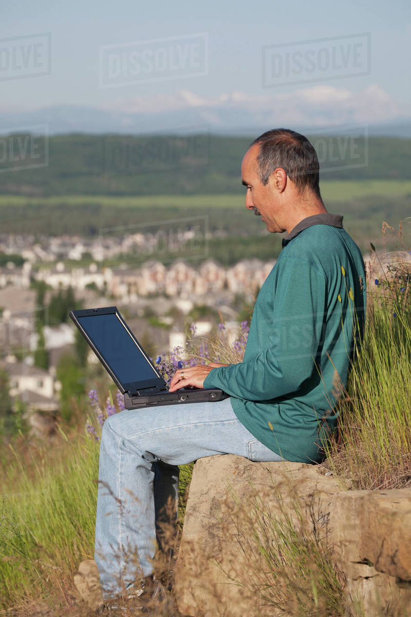 Man Typing On A Laptop Computer Sitting On A Rock Ledge Overlooking A ...