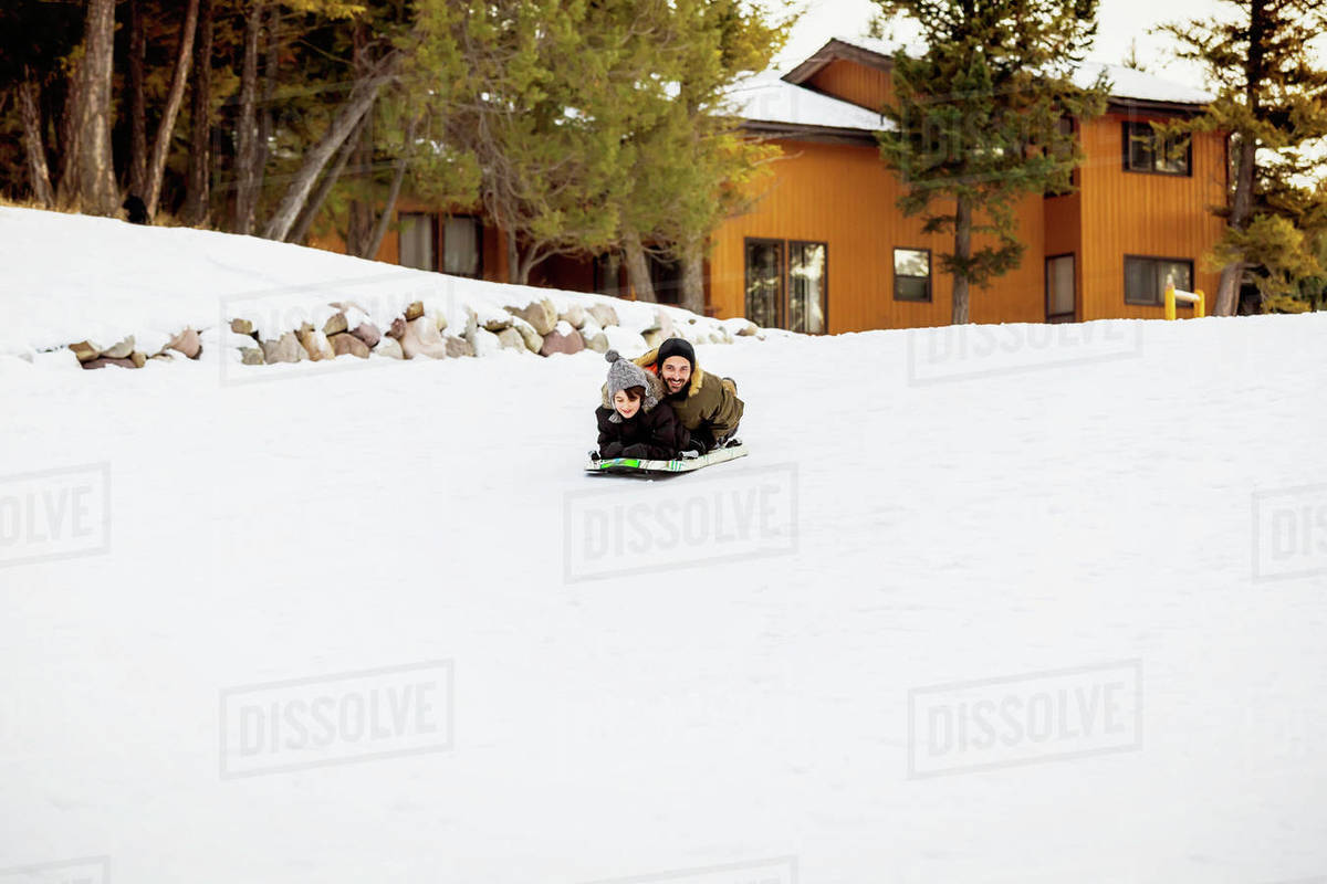 Father And Son Ride Down A Snowy Hill Together Laying Down On A Sled At ...