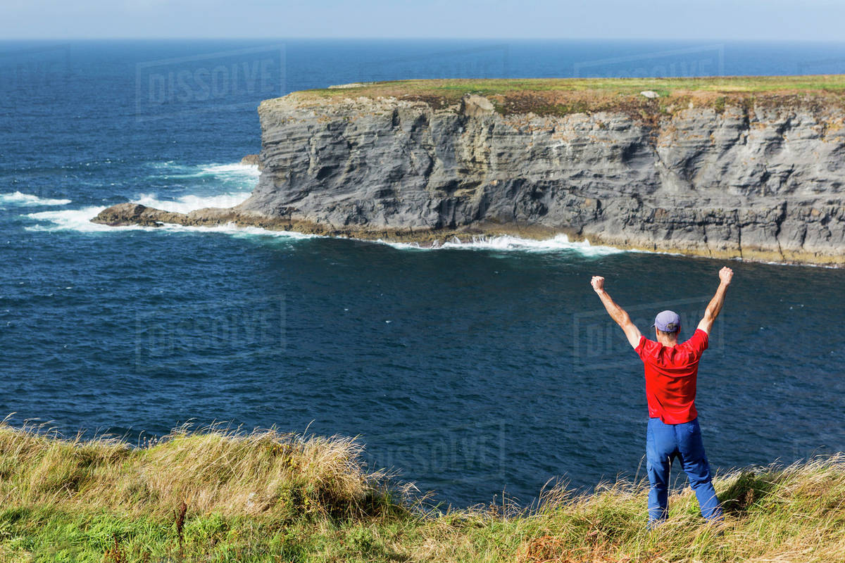 Man standing on grassy cliff's edge with arms raised overlooking large ...