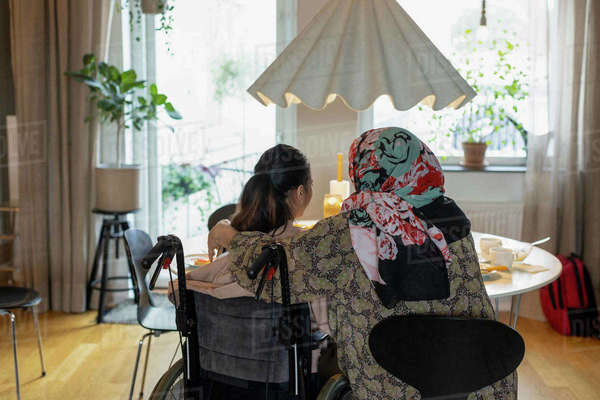 Rear view of mother sitting with daughter having paraplegia at home ...
