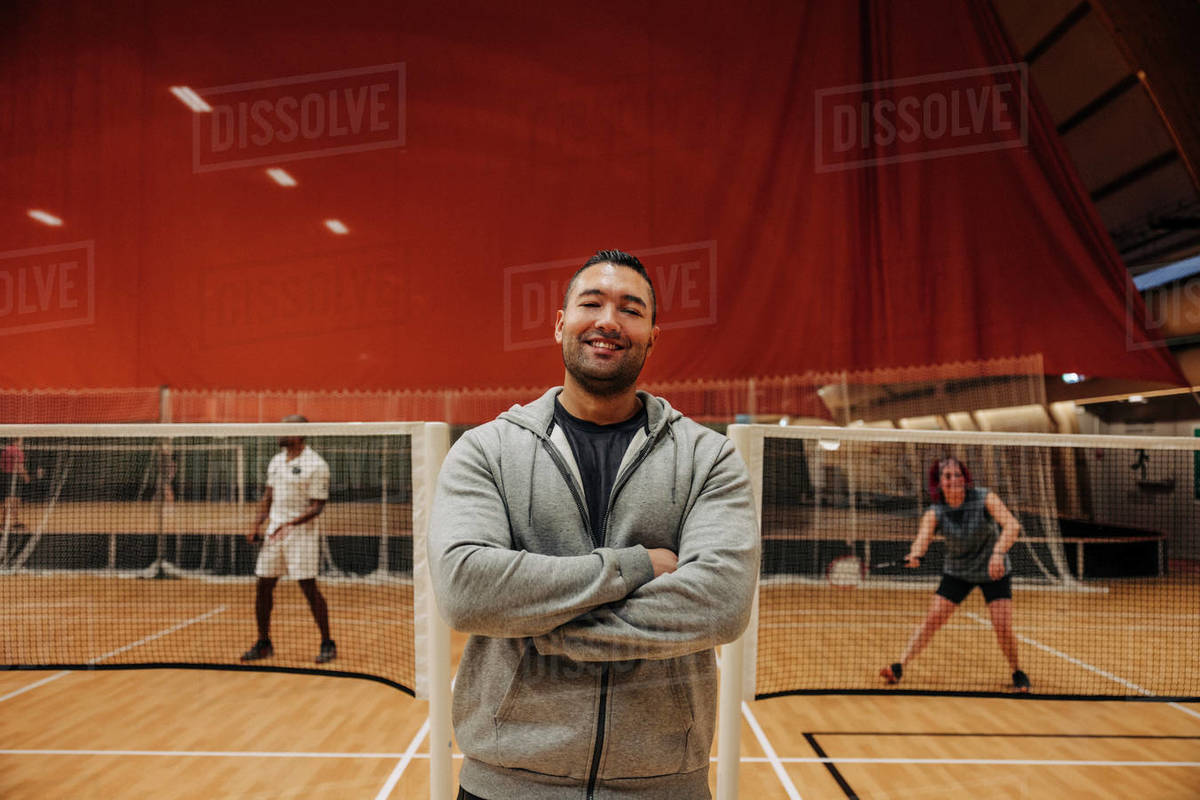 Smiling male coach standing with arms crossed at badminton court ...