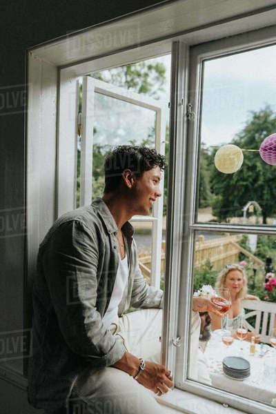 Smiling young man sitting on window sill while looking at friends ...
