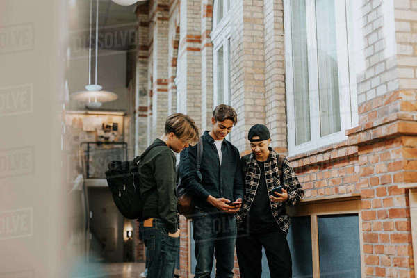 Teenage boy sharing mobile phone with friends while walking in school ...