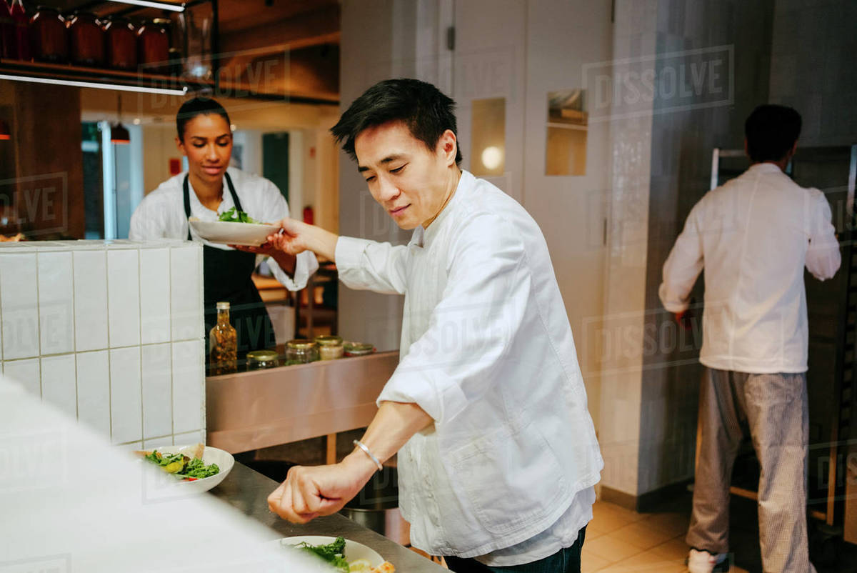 Male chef passing salad bowl to female coworker while finishing salad ...