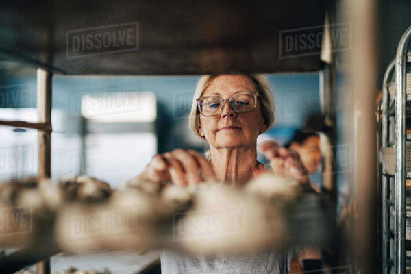 Female senior baker arranging buns on cooling rack while working at ...