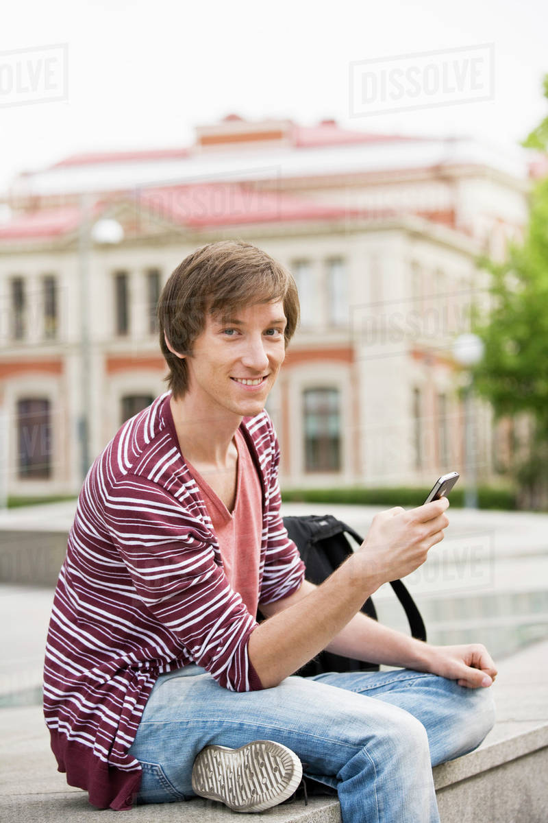 Portrait of happy male university student with cell phone sitting at ...