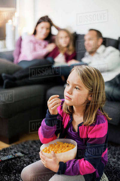 Girl having snacks while watching TV on floor with family in background ...
