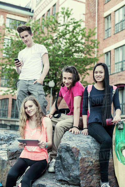 Portrait of smiling high school students sitting on rock at schoolyard ...