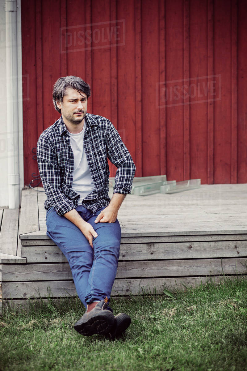 Full length of thoughtful male farmer sitting on porch at yard ...