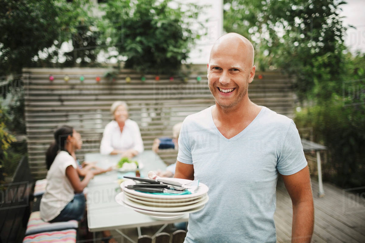 Portrait of happy man holding plates with family sitting at outdoor ...