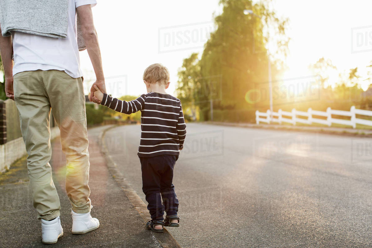 Rear view of boy walking with father while holding hands on street ...