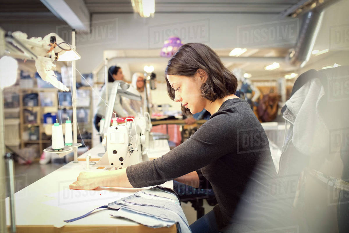 Side view of woman sewing fabric on machine while volunteers working in ...