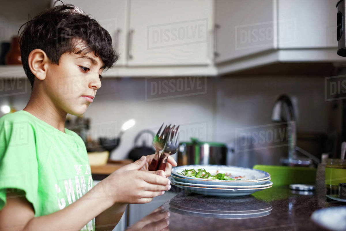 Side view of boy holding forks while standing at kitchen counter ...
