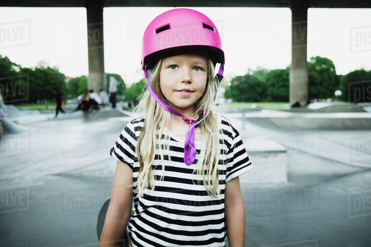 Portrait of smiling girl wearing pink helmet standing at skateboard ...