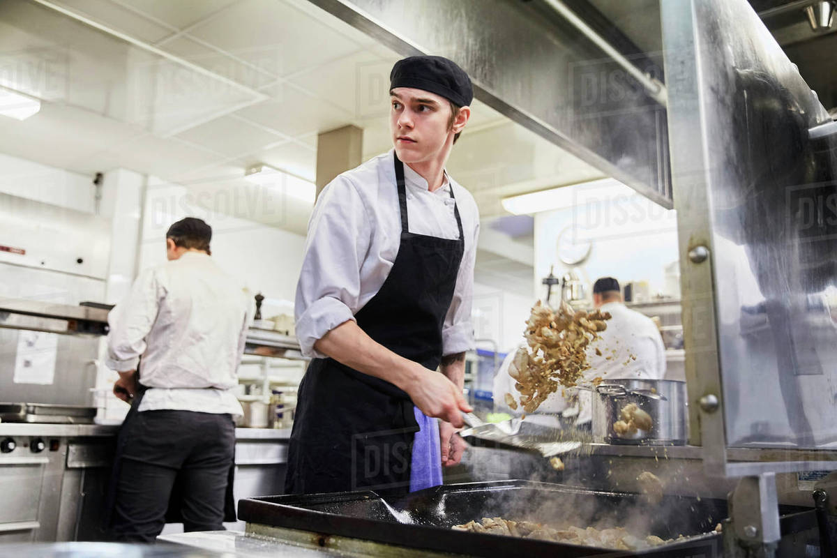Male chef student tossing meat with colleagues in background at cooking ...