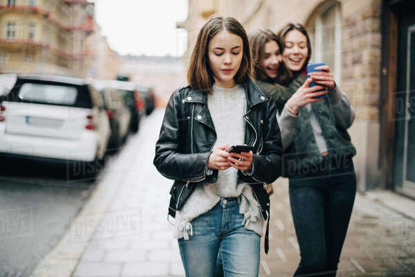 Female friends using mobile phone while walking on sidewalk in city ...