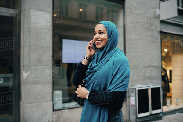 Happy young Muslim woman talking on mobile phone walking building in ...
