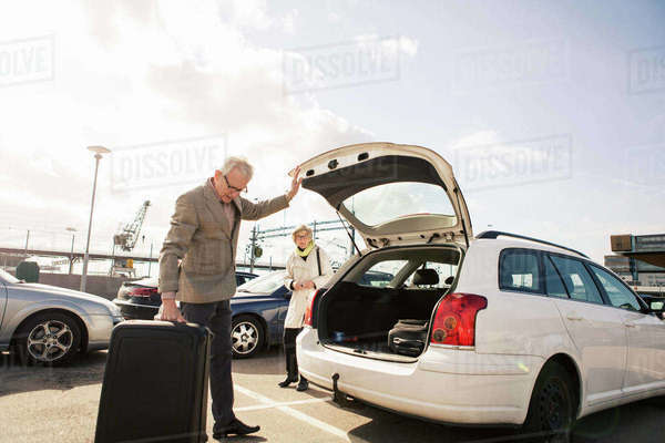 Senior man loading luggage in car trunk with woman standing in parking ...